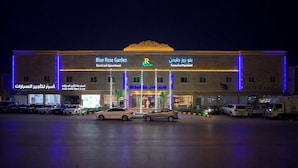 Hotel com entrada de serviço, vários carros estacionados e sinalização iluminada.