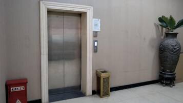 Um elevador com porta, uma pequena lixeira, uma lixeira vermelha e um vaso decorativo grande no chão.