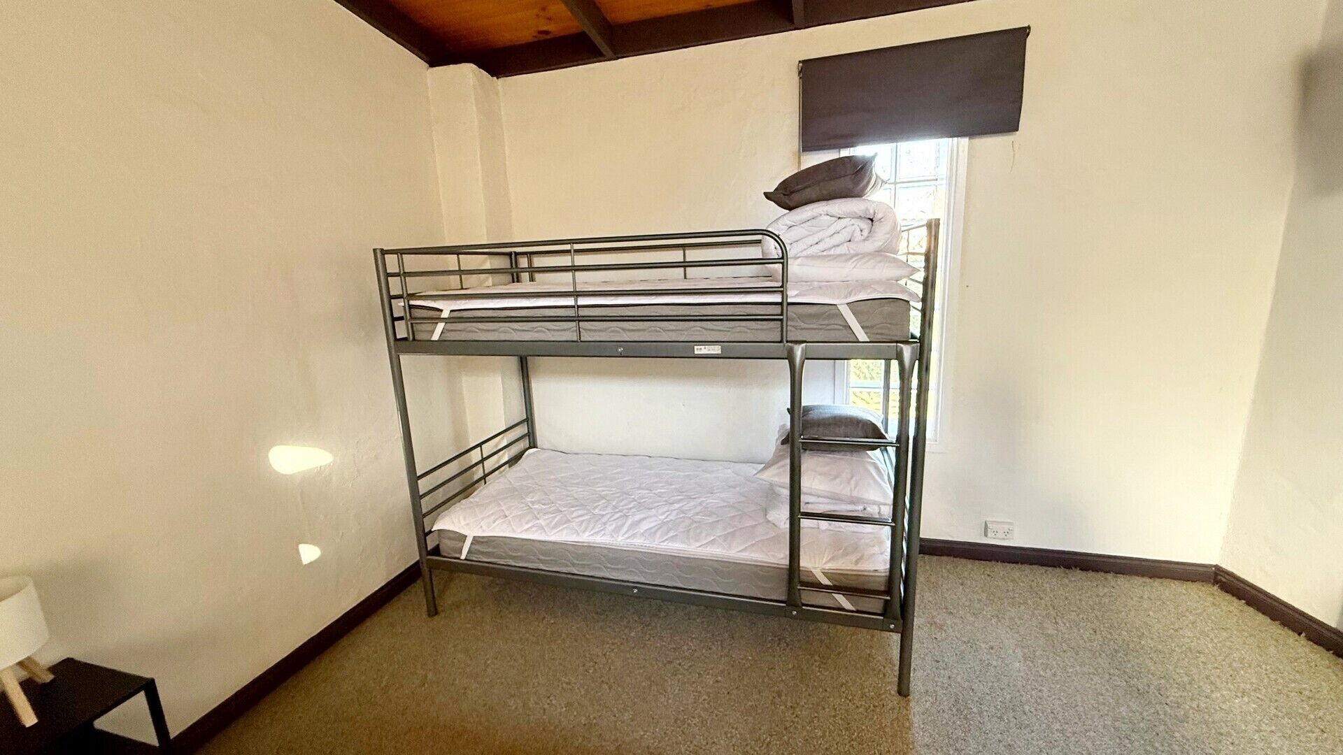 Quarto com beliche, janela, chão coberto de carpete e uma mesinha com luminária.