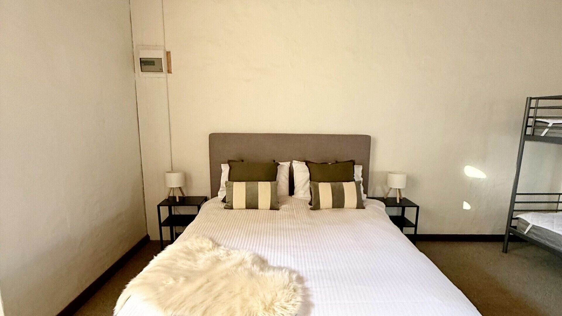 Uma cama de solteiro com um cobertor branco fofo, duas mesas de cabeceira com abajures, uma cama beliche à direita e uma parede lisa com um termostato.