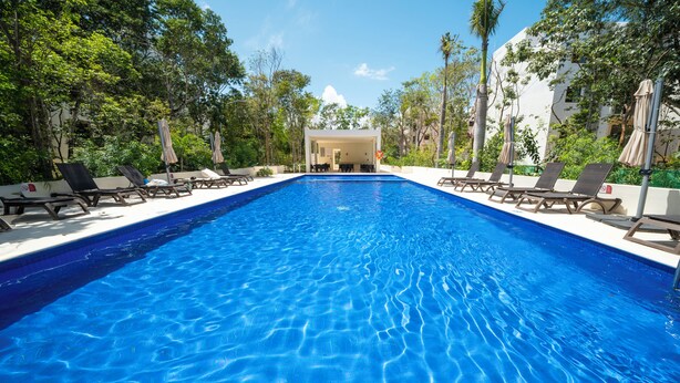 Piscina azul límpida com espreguiçadeiras e guarda-sóis, rodeada por árvores e um edifício ao fundo.