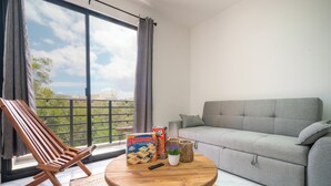 Uma sala de estar moderna com um sofá cinza, uma cadeira de madeira, uma mesa redonda pequena com um jogo de tabuleiro, um controle remoto e um vaso com planta.