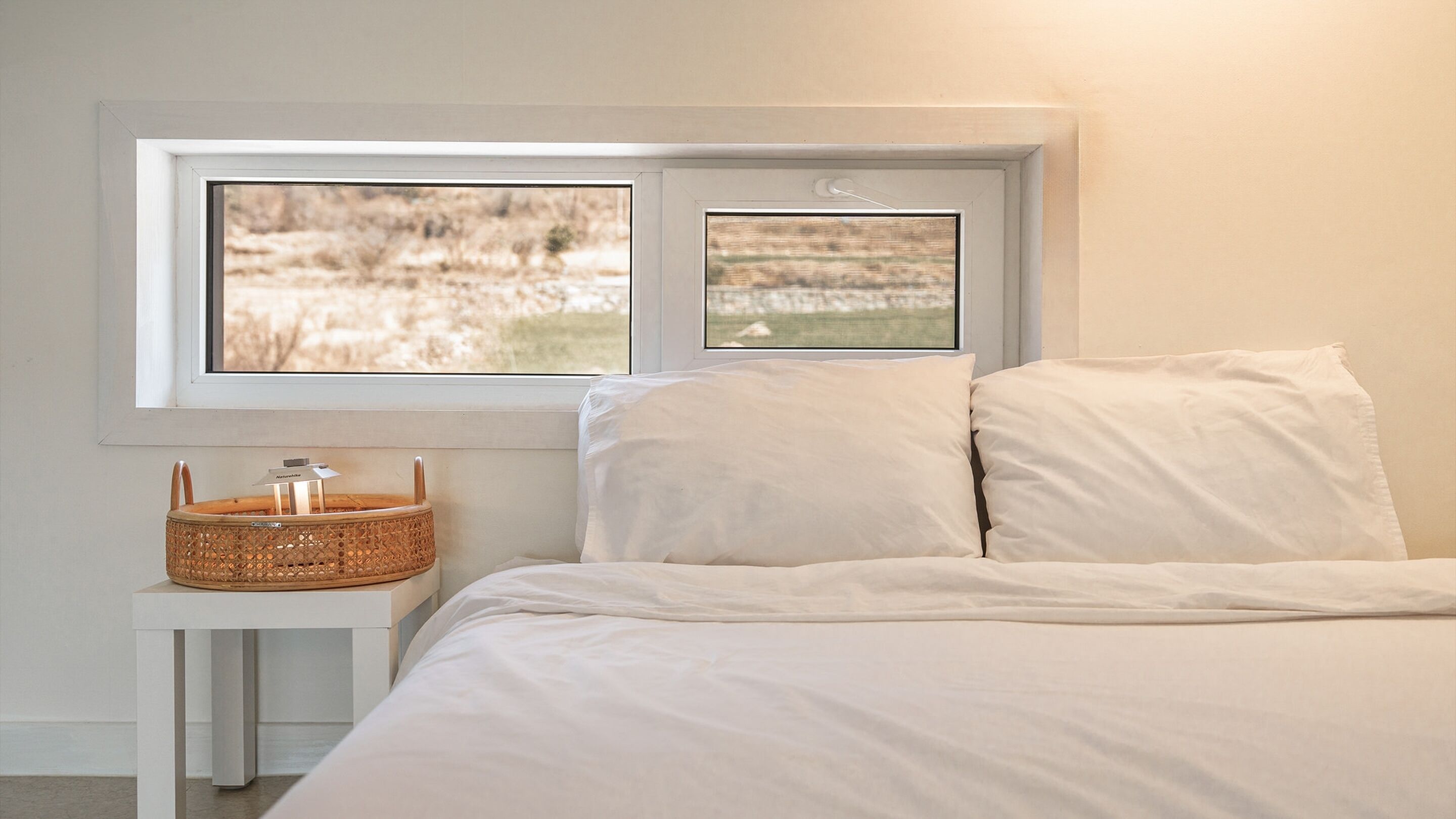 Quarto com cama, almofadas brancas, mesa de cabeceira com um cesto e janela com vista para um campo.
