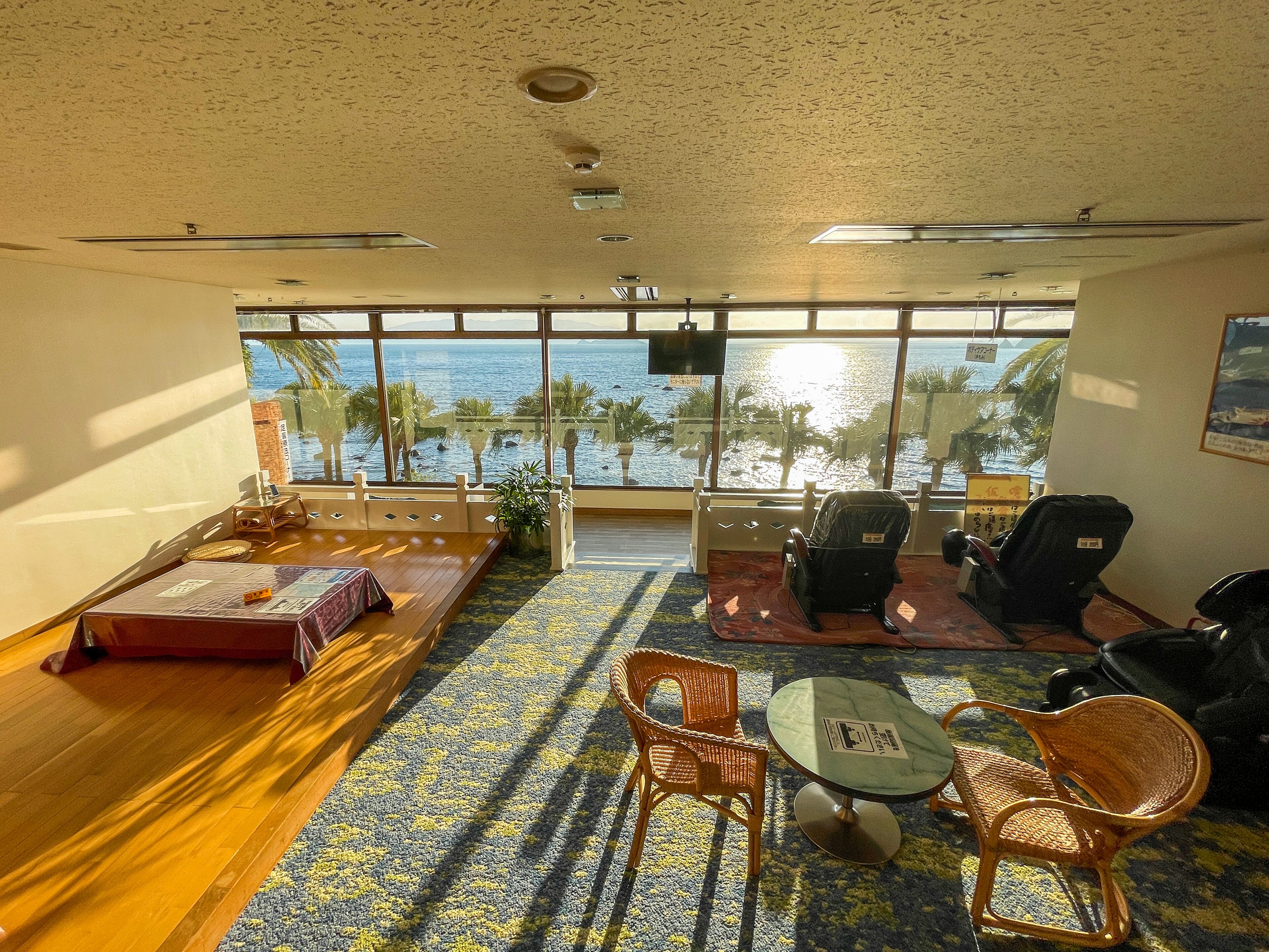 Quarto com uma grande janela com vista para a praia, piso de madeira, uma mesa pequena e algumas cadeiras.