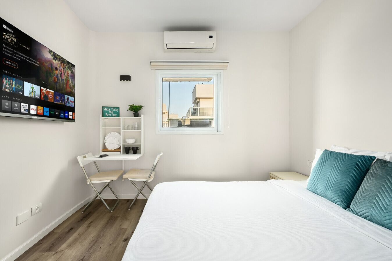 Quarto de hotel com cama, escrivaninha com cadeira, janela e televisão na parede exibindo um filme.