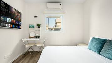 Quarto de hotel com cama, escrivaninha com cadeira, janela e televisão na parede exibindo um filme.
