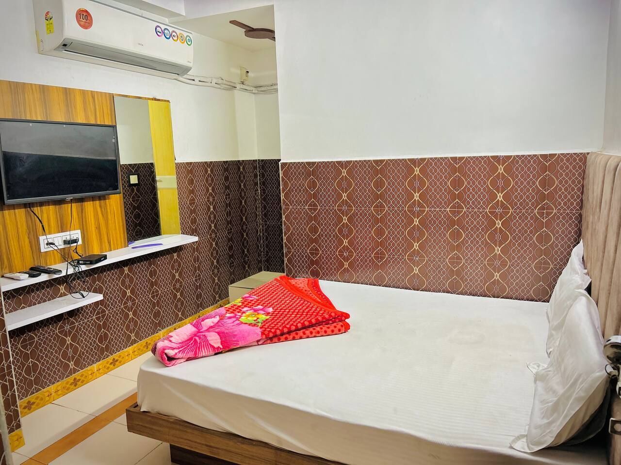 Quarto de hotel com uma cama, uma TV fixada na parede, uma prateleira pequena com objetos e um papel de parede estampado.
