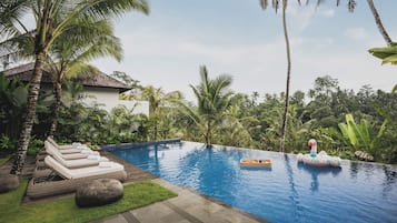 Piscina com espreguiçadeiras e plataforma flutuante, cercada por vegetação tropical.