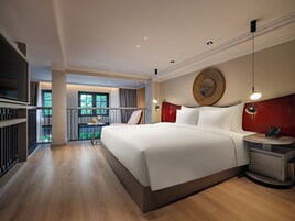 Quarto de hotel moderno com uma cama grande, uma mesa de cabeceira com telefone, um espelho redondo e vista para uma varanda com grade.