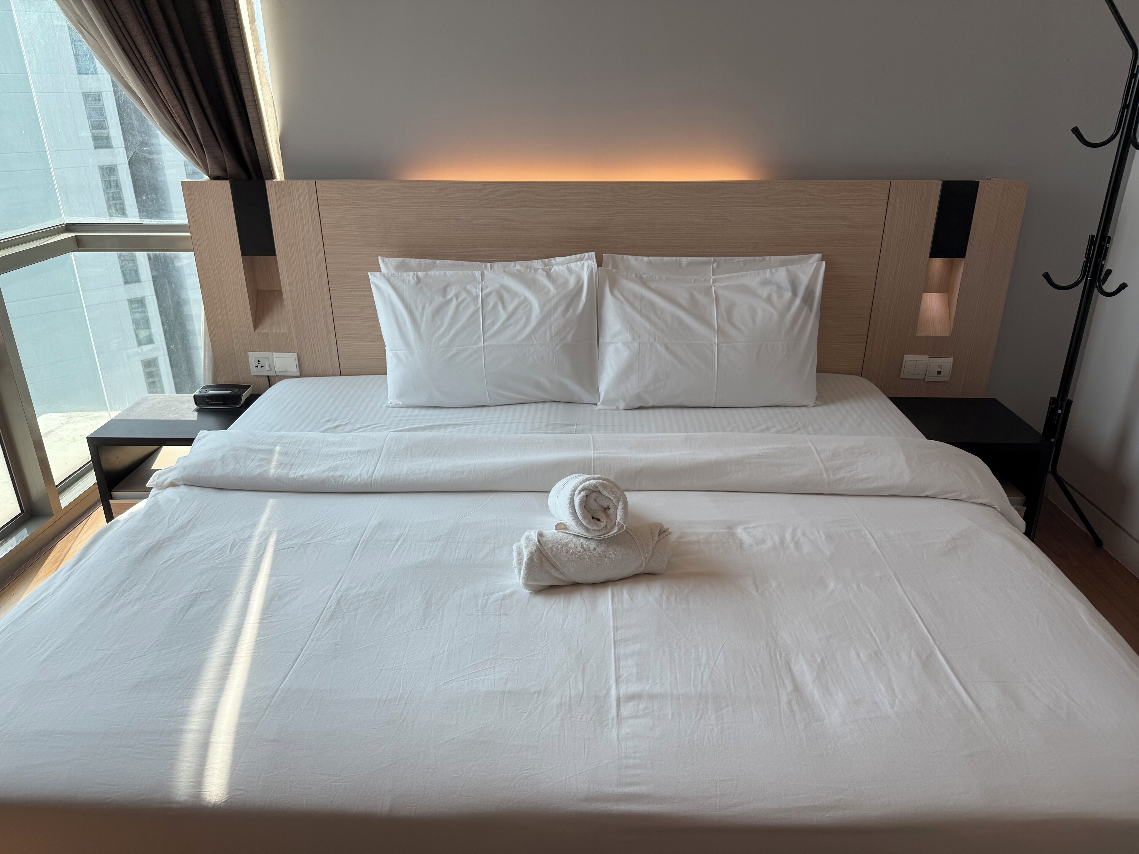 Cama bem arrumada com lençóis brancos, toalha dobrada e mesa de cabeceira com abajur.