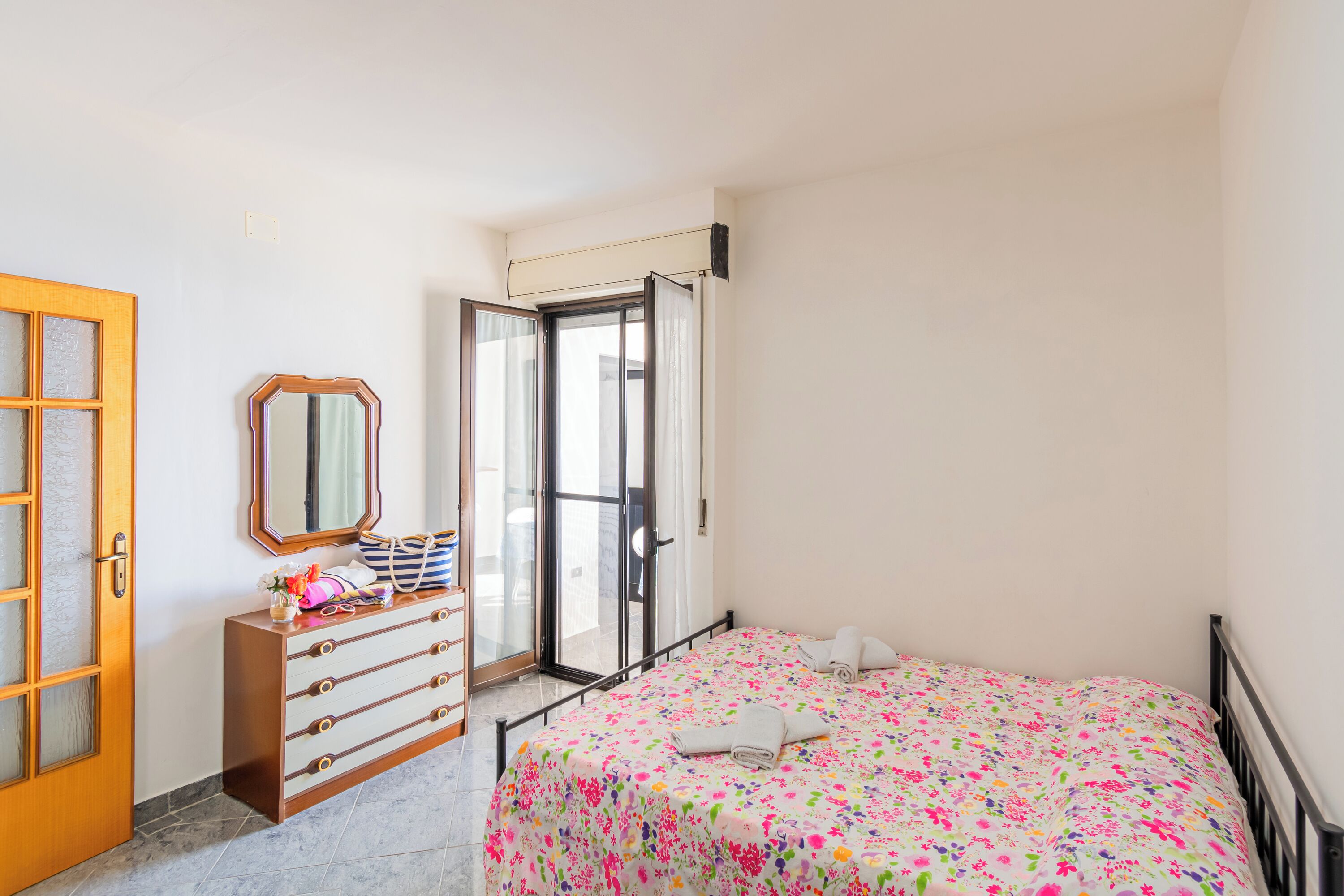 Um quarto com cama, cômoda, espelho e porta que dá para uma varanda.