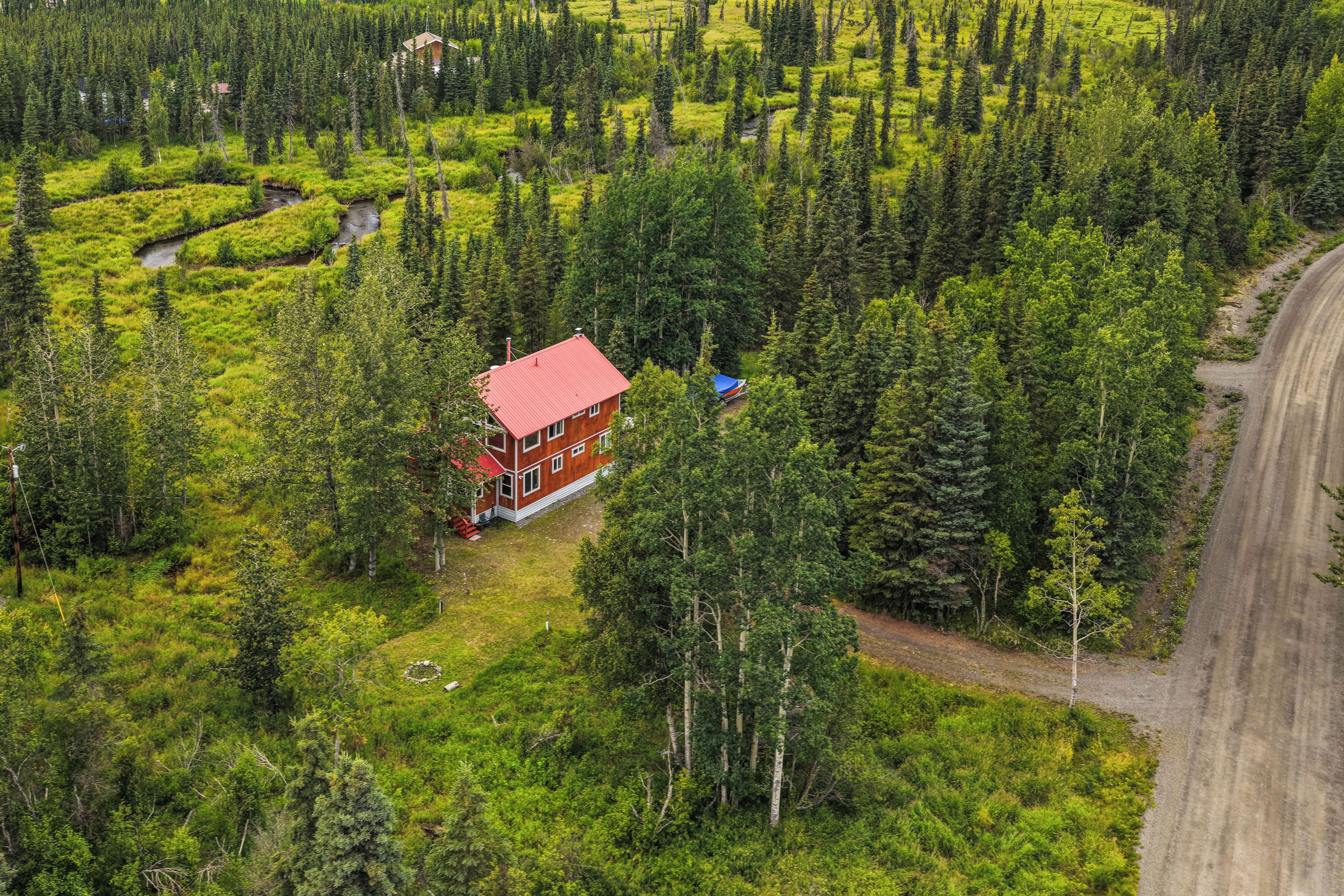 Uma casa de dois andares com telhado vermelho, cercada por uma floresta densa e um pequeno riacho.