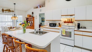 Uma cozinha moderna com armários brancos, uma ilha central com pia e uma área de jantar com cadeiras de vime.