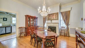 Uma sala de jantar com mesa de madeira, cadeiras, um candelabro, lareira e cozinha visível ao fundo.
