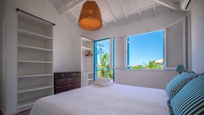 Quarto com uma cama grande, guarda-roupa, luminária pendente e janela com vista para palmeiras.