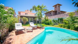 Piscina com espreguiçadeiras, rodeada por vegetação exuberante e uma casa grande com uma torre.