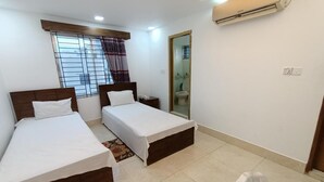 Quarto de hotel com duas camas de solteiro, uma janela com cortinas, unidade de ar condicionado e casa de banho visível através de uma porta de vidro.