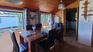 Uma área de refeições em madeira com uma mesa posta para quatro pessoas, uma cozinha ao fundo e vista para uma casa móvel lá fora.