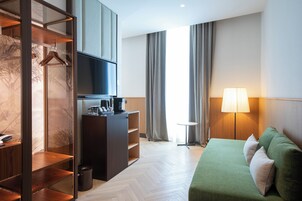 Quarto de hotel com sofá verde, televisão, mesa pequena com abajur e guarda-roupa com espelho.