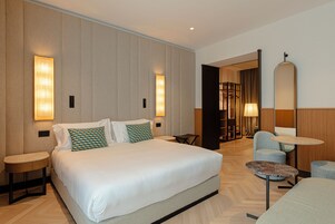 Quarto de hotel moderno com cama grande, duas mesas de cabeceira, uma mesa de centro redonda e um espaço de estar com poltrona e luminária.