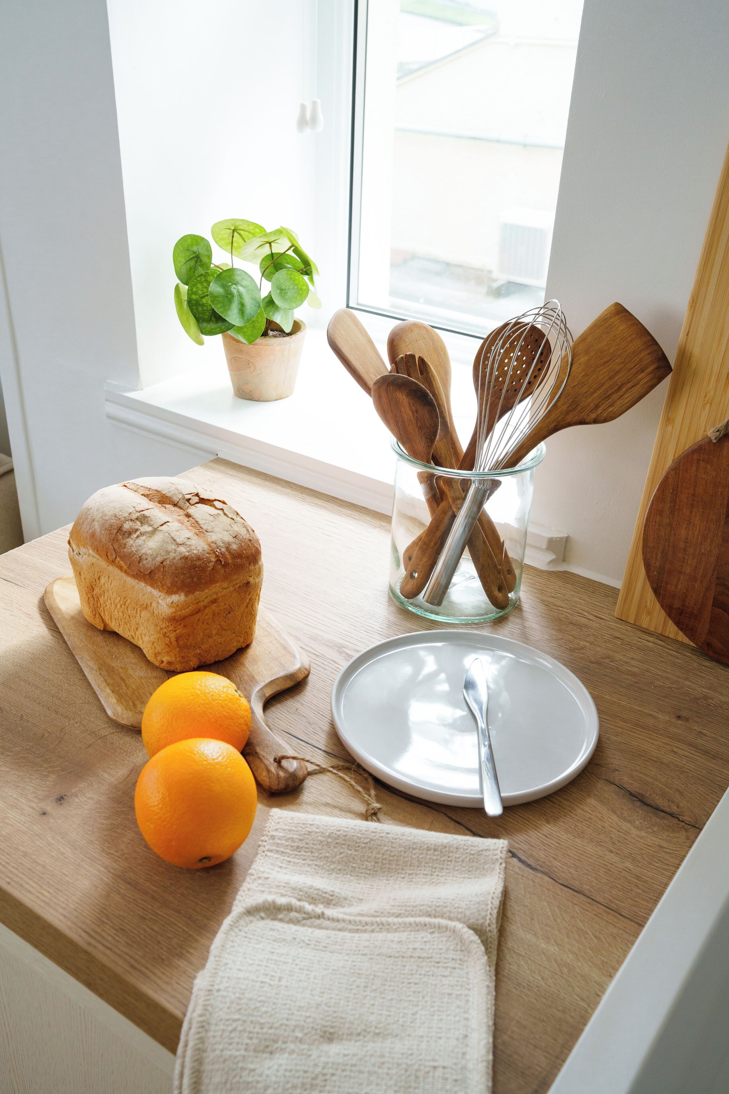 Uma bancada de cozinha com um pão, laranjas, uma tábua de corte, um prato, um conjunto de batedores e uma planta em vaso.
