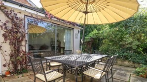 Um pátio externo com mesa e cadeiras de metal, um guarda-sol amarelo e uma porta de vidro que dá para um espaço interno.