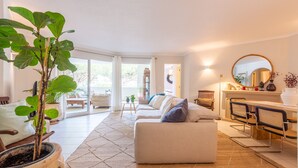 Uma sala de estar espaçosa com sofá, mesa de centro e área de jantar com cadeiras. Há uma janela grande com cortinas, lareira e um espelho na parede.