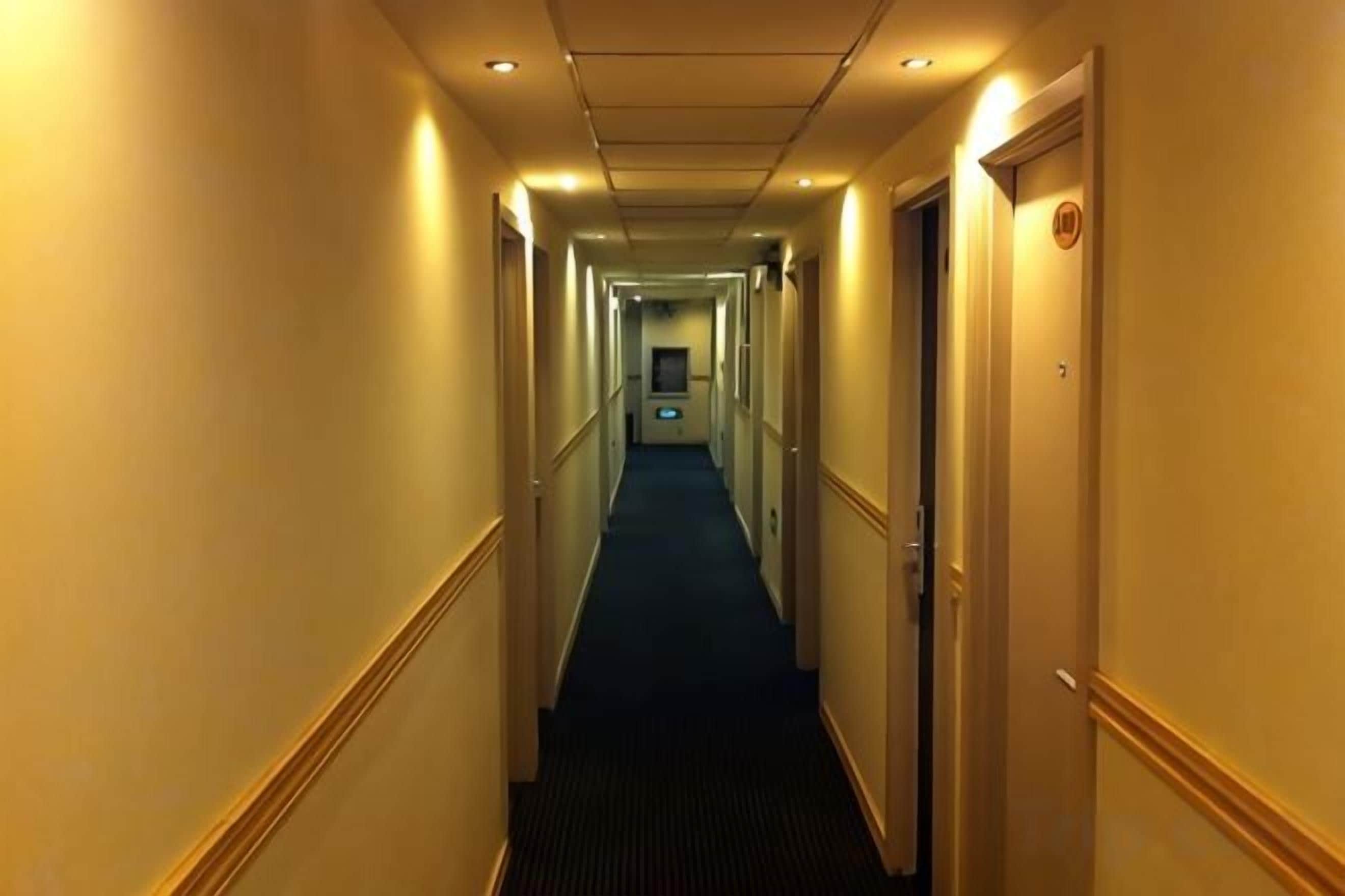 Corredor de hotel com portas em ambos os lados, piso coberto de carpete e teto com iluminação embutida.