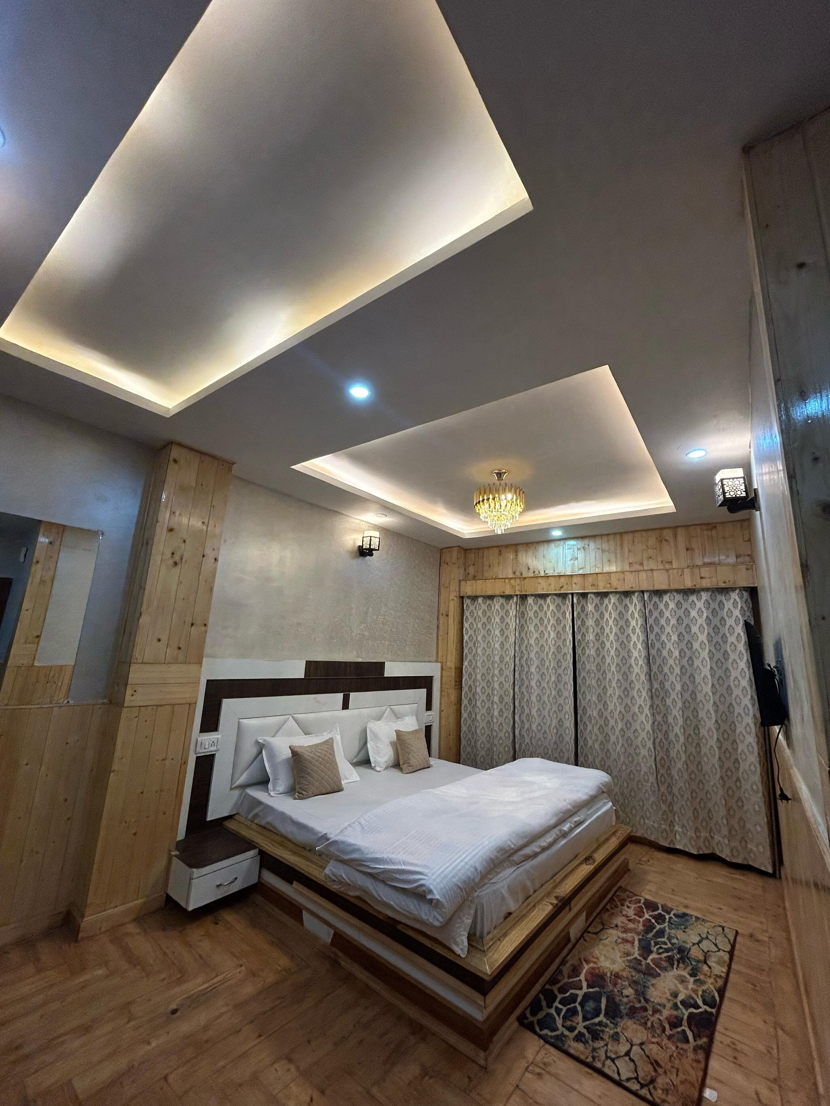 Quarto moderno com cabeceira de madeira, uma cama grande com roupa de cama branca, um candelabro e iluminação embutida no teto.