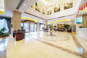 Um saguão de hotel com balcão de recepção, uma fonte decorativa central e um grande candelabro.
