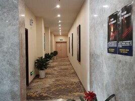 Corredor de hotel com piso estampado, plantas em vasos, quadros nas paredes e um cartaz divulgando um evento do hotel.