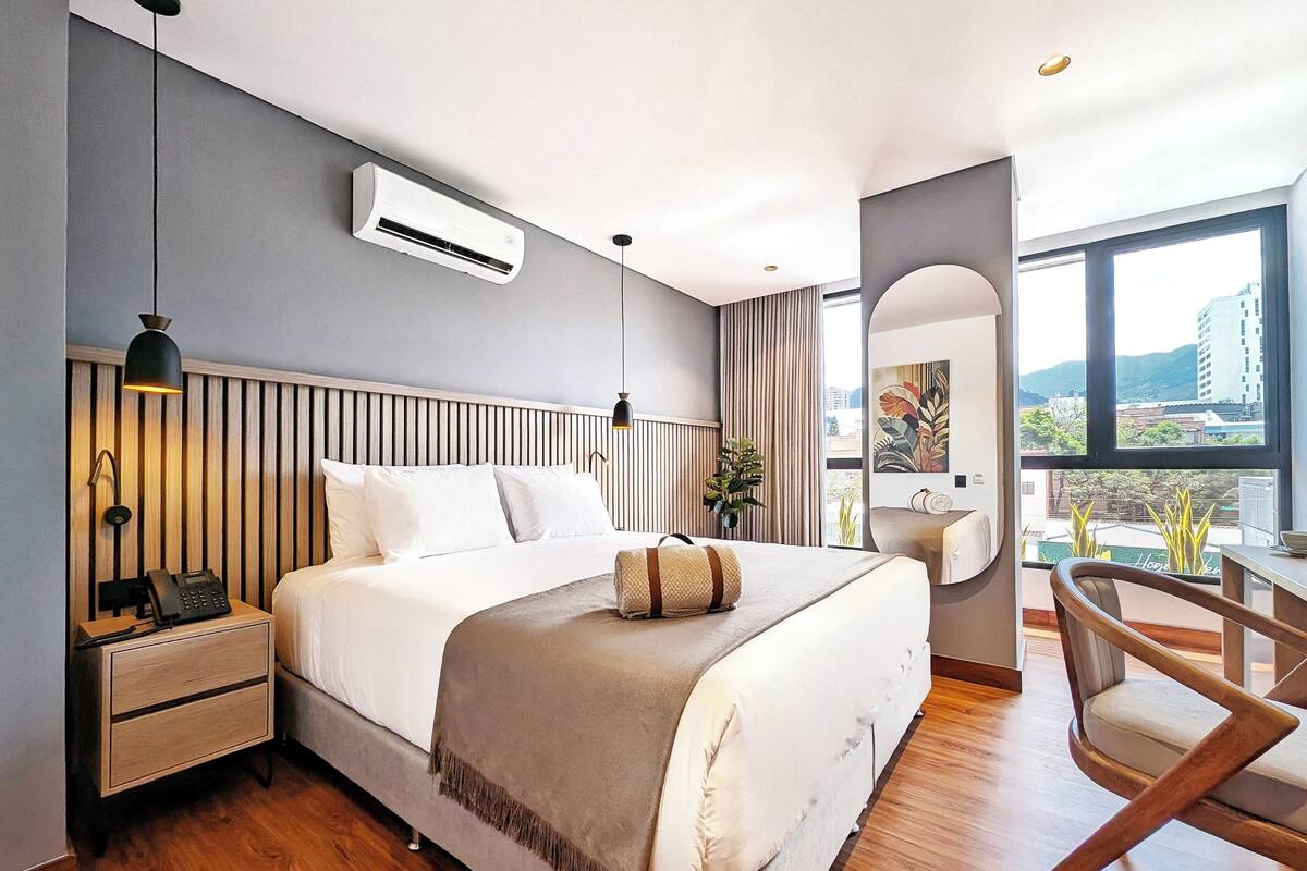 Quarto de hotel moderno com uma cama grande, mesas de cabeceira, uma escrivaninha e vista para a cidade.