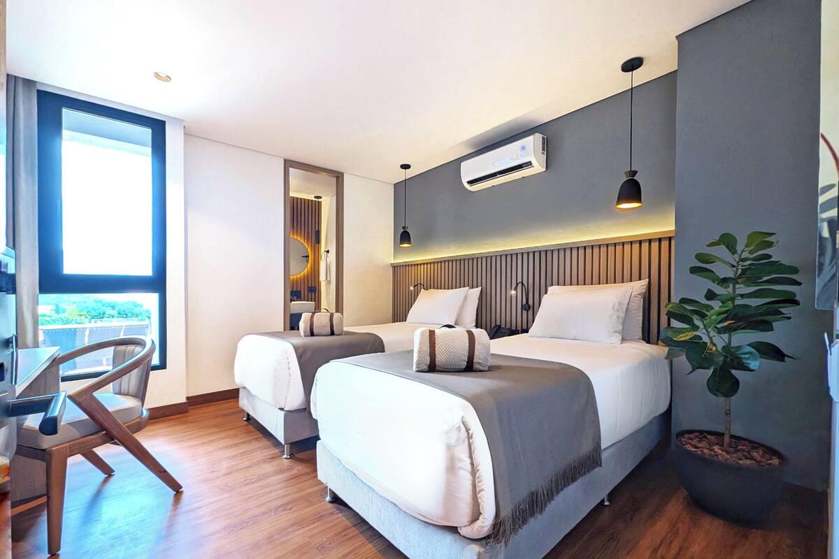 Quarto de hotel moderno com duas camas, uma escrivaninha e uma janela grande.