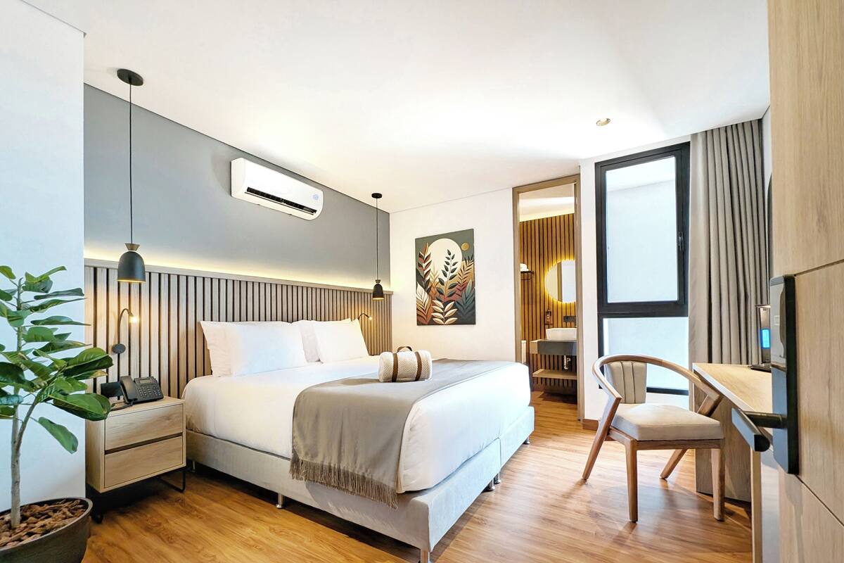 Quarto de hotel moderno com uma cama grande, mesa de cabeceira com luminária, escrivaninha com cadeira e uma planta em vaso.