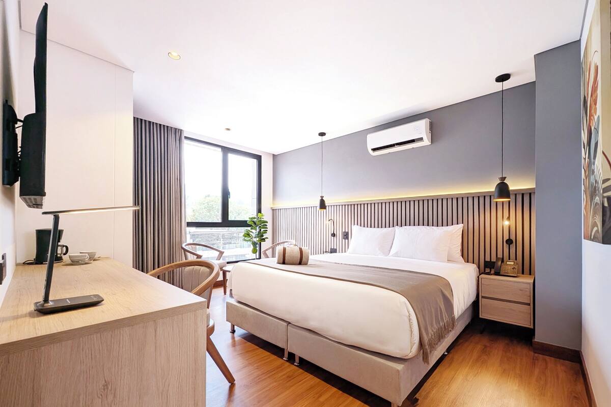 Quarto de hotel moderno com cama, mesa de cabeceira, luminária, aparelho de ar condicionado e vista para o exterior.