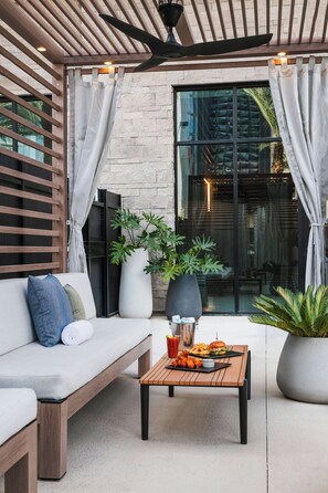Uma área externa com mesa e cadeiras, comida, plantas em vasos e vista para o interior do edifício através de uma porta de vidro.