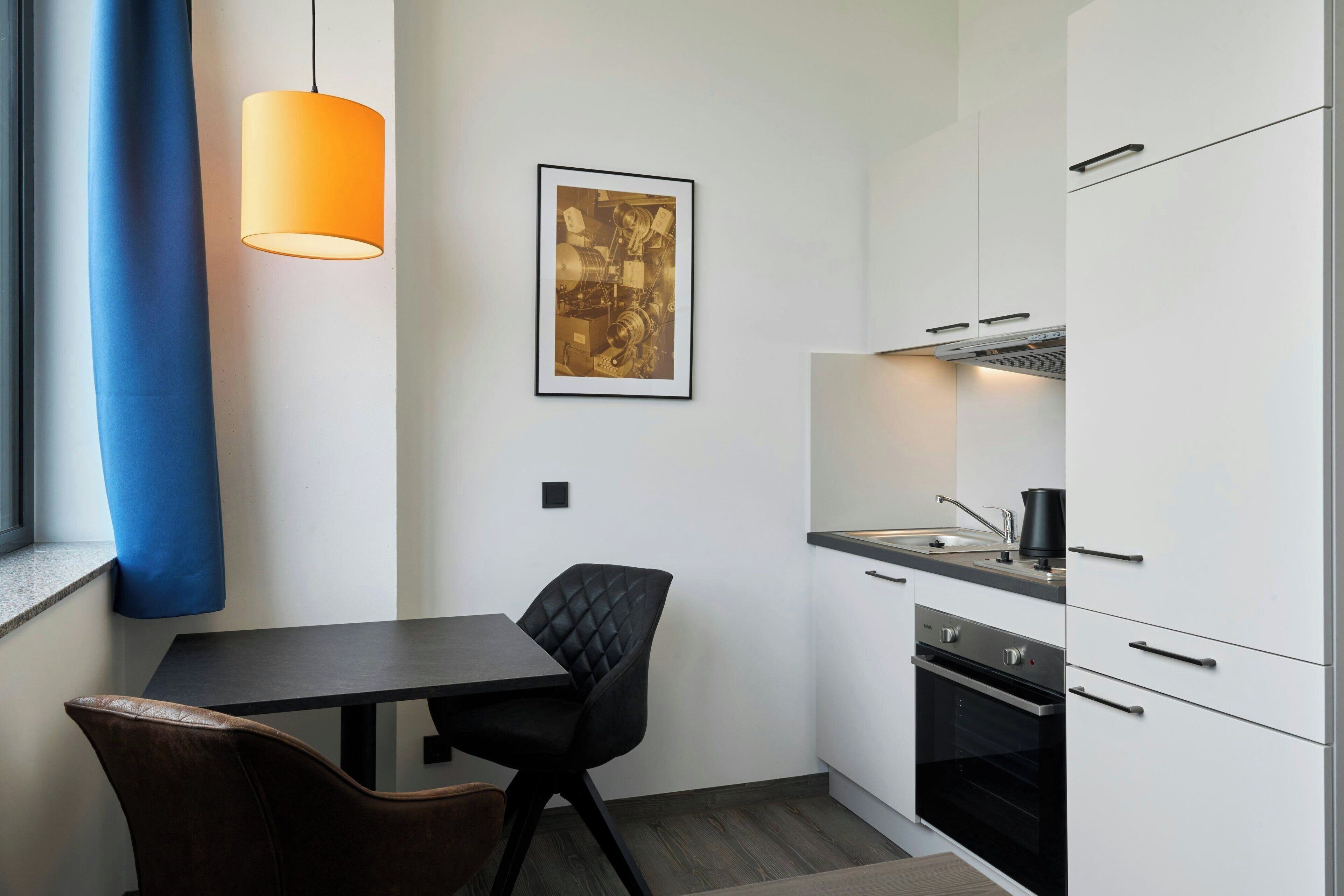 Uma cozinha moderna com armários brancos, bancada preta e forno embutido. Há um quadro na parede e uma luminária pendente sobre a mesa.