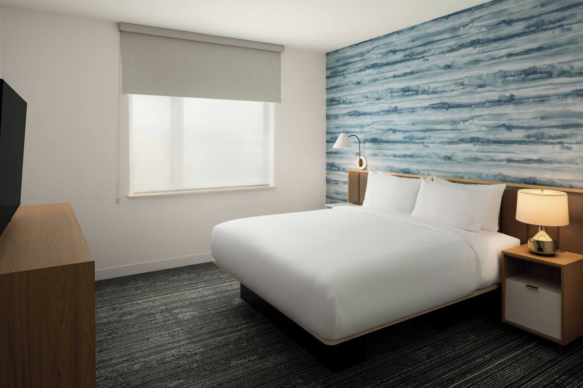 Quarto de hotel com uma cama grande, uma mesa de cabeceira com abajur, uma escrivaninha de madeira, uma janela com persiana e uma parede texturizada.