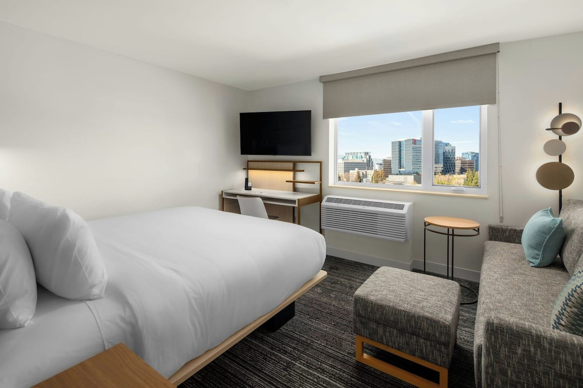 Quarto de hotel com cama, escrivaninha com cadeira, sofá, televisão na parede e janela com vista para a cidade.