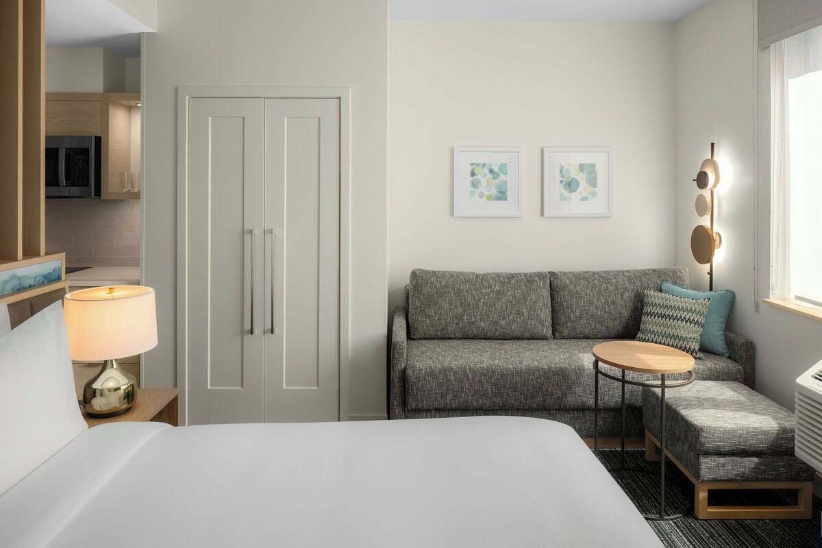 Quarto de hotel com cama, sofá, mesa redonda pequena, luminária, porta e duas imagens emolduradas na parede.