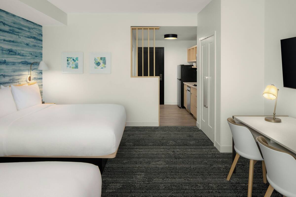 Quarto de hotel com uma cama grande, uma mesa com duas cadeiras, uma kitchenette e obras de arte nas paredes.