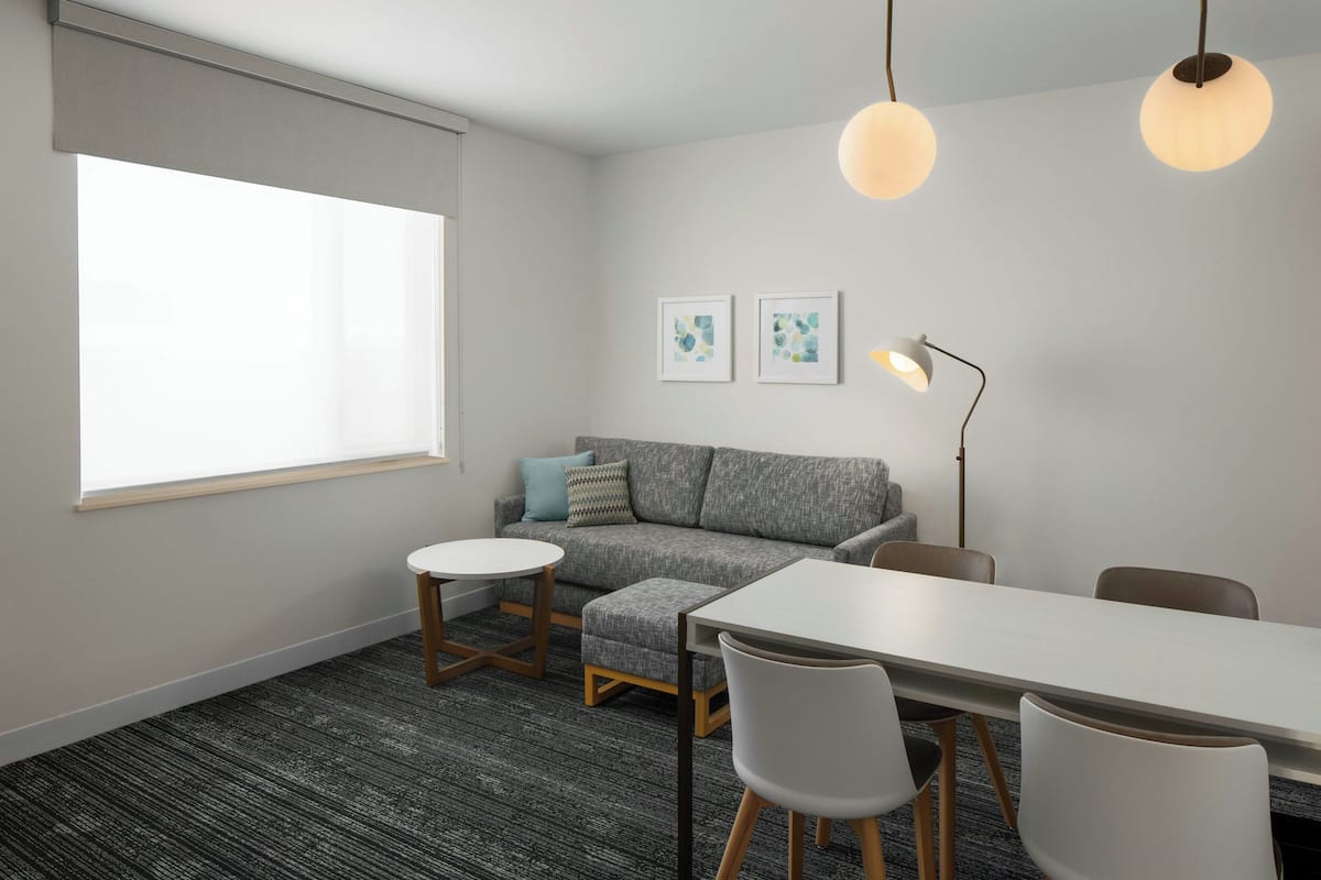 Quarto moderno com sofá cinzento, mesa redonda e longa mesa de jantar. Há duas luzes pendentes, um abajur e duas fotografias emolduradas na parede.