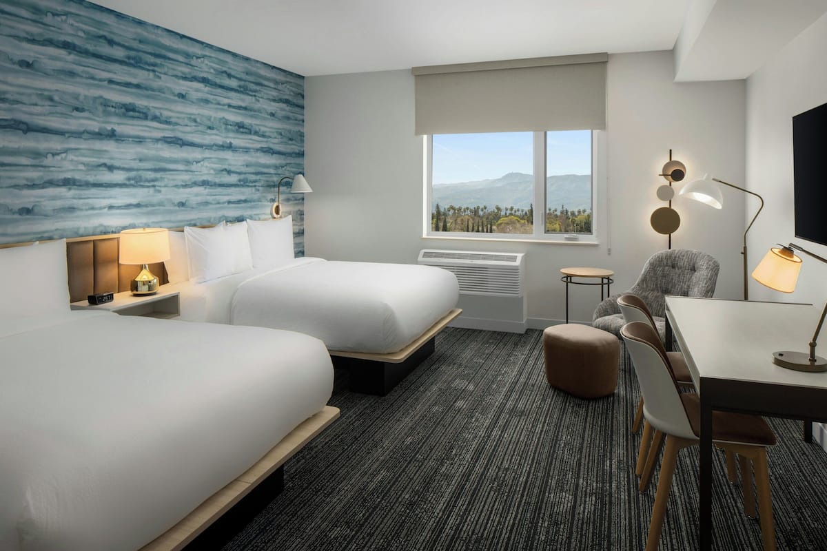 Quarto de hotel com duas camas, uma escrivaninha, uma cadeira, uma mesinha, um abajur, TV na parede e uma janela com vista para as montanhas.