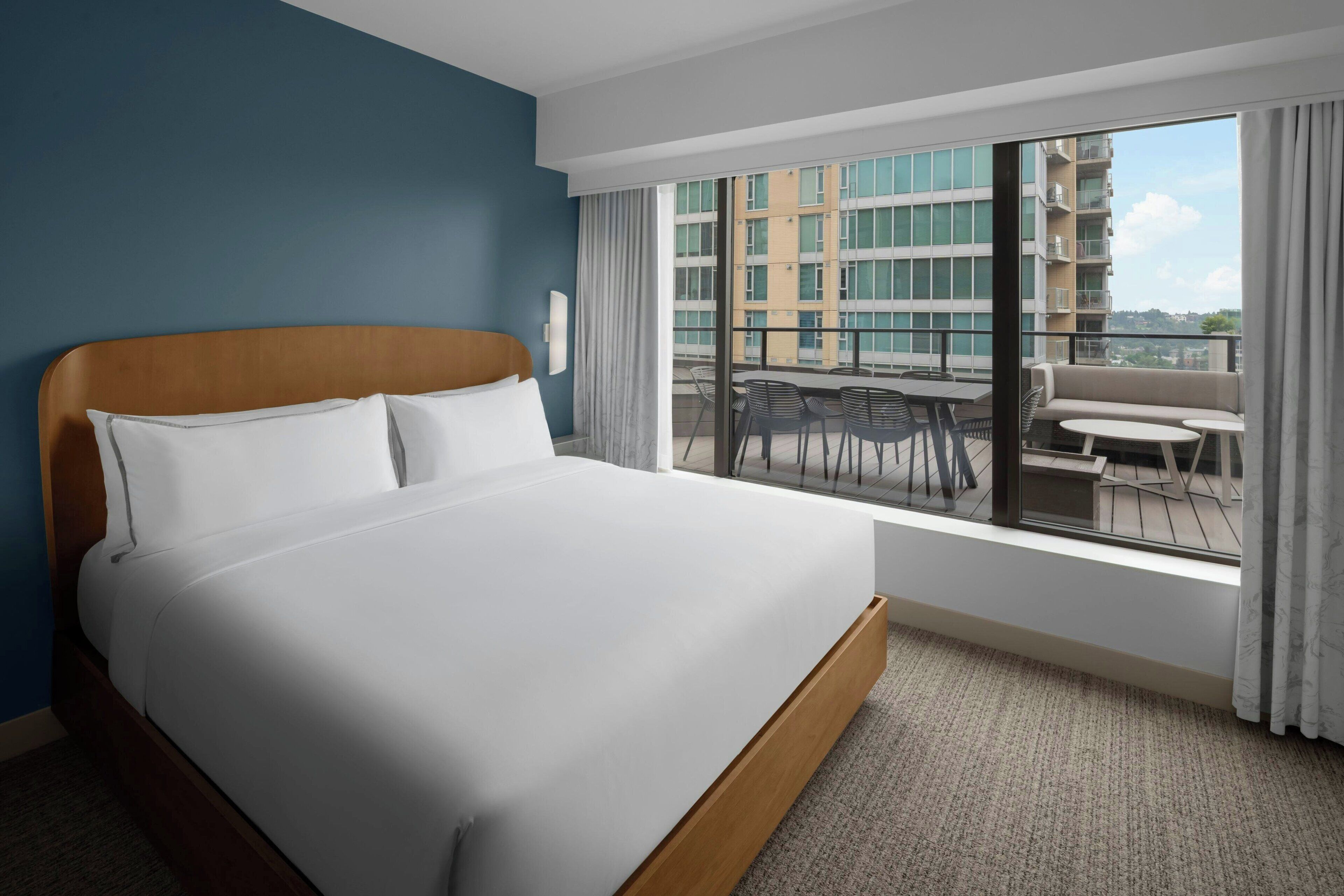 Quarto de hotel com uma cama grande, vista da cidade através de uma varanda e área de estar com mesas e cadeiras.