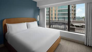 Quarto de hotel com uma cama grande, vista da cidade através de uma varanda e área de estar com mesas e cadeiras.