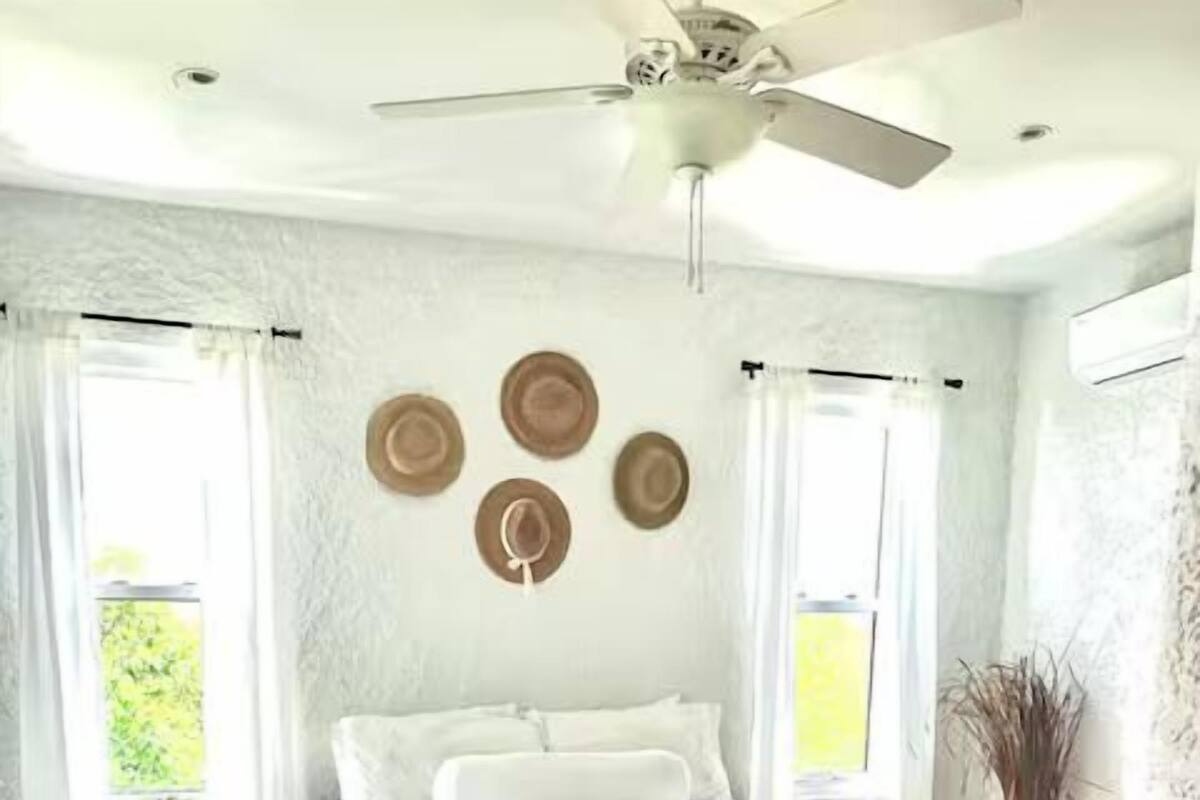 Quarto com estrutura de cama de madeira, roupa de cama branca, ventilador de teto e uma pequena mesa com uma planta.