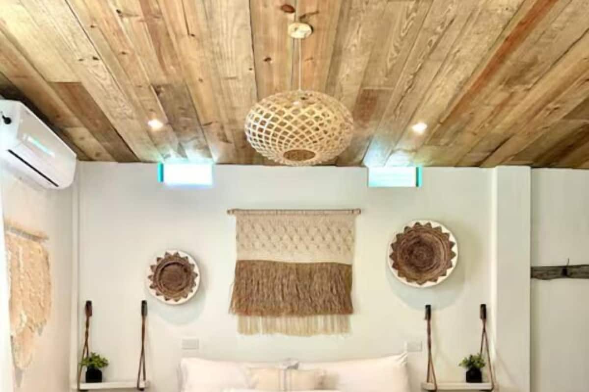 Quarto com cabeceira de madeira, colcha branca e dois objetos decorativos fixados na parede.
