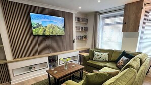 Uma sala de estar moderna com um sofá verde, uma mesa de centro de madeira, uma TV de tela plana presa a uma parede com design em painel de madeira, e uma estante cheia de livros e objetos decorativos.
