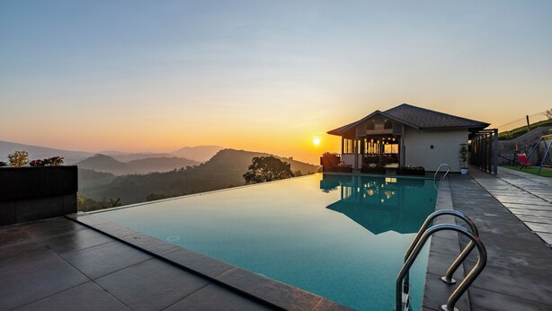 Piscina com vista para montanhas ao pôr do sol, área de estar coberta e um pátio com uma cadeira.