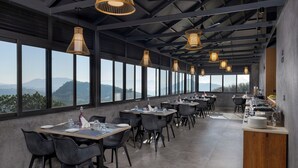 Ampla sala de jantar com pé direito alto e grandes janelas com vista para as montanhas. Mesas postas com pratos e talheres, e vários lustres.
